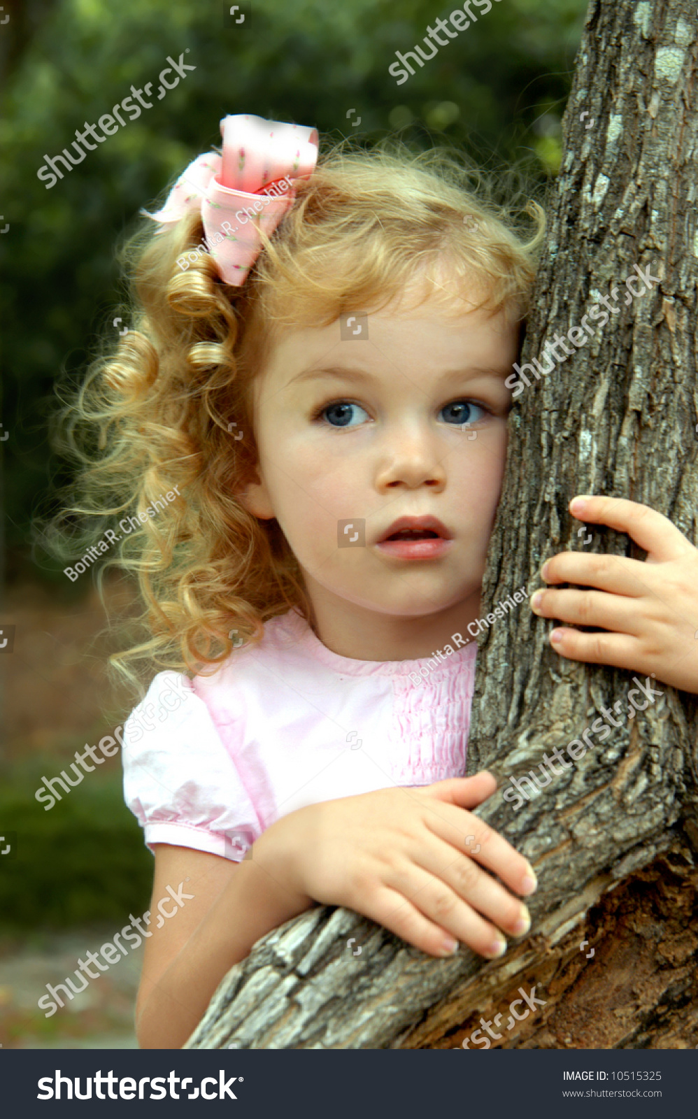Young Girl Peeks Out From Behind A Tree She Has Golden Curls And A