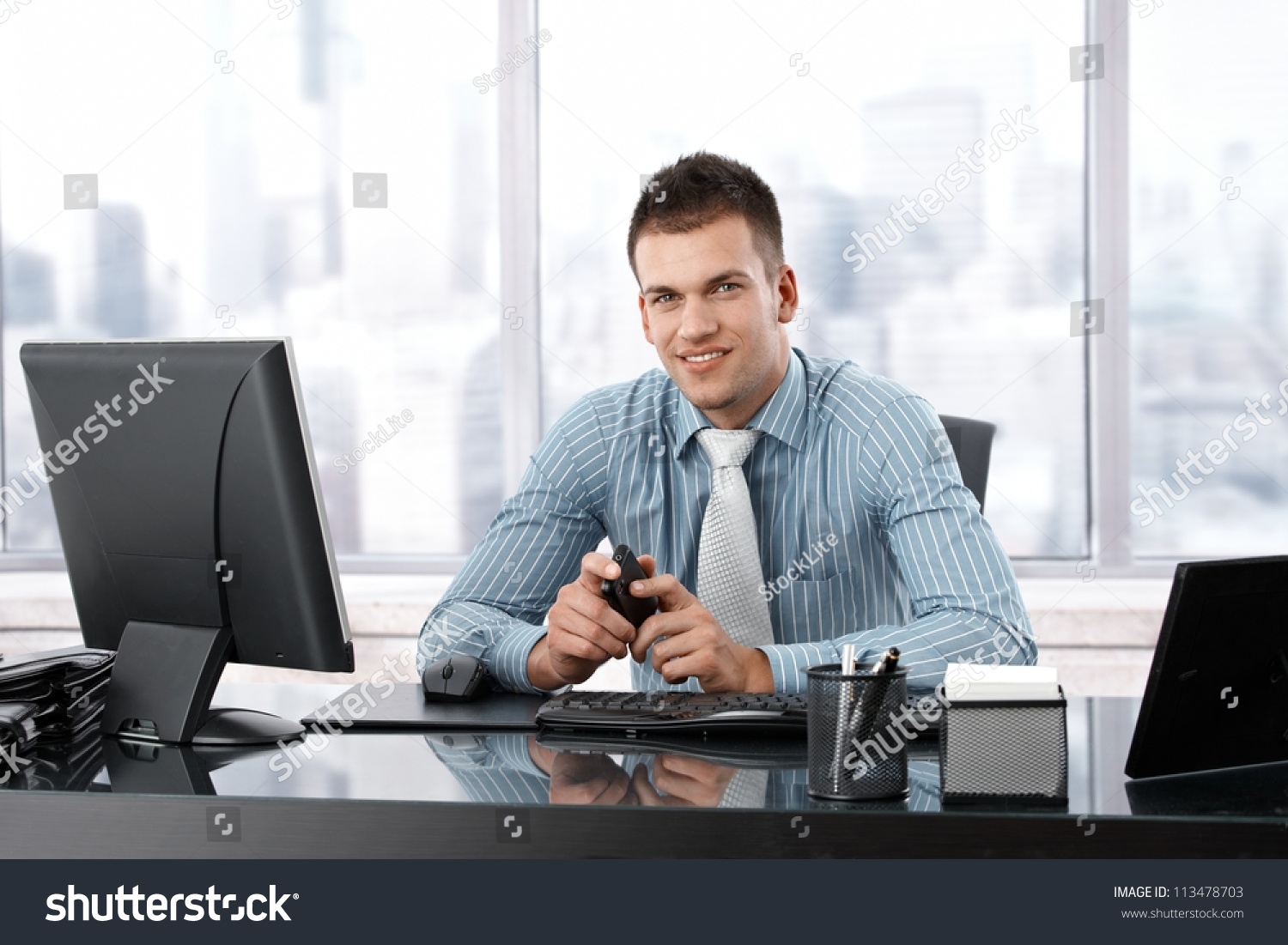 Young Manager Sitting At Desk In Bright Office, Smiling Confidently ... Young Manager Sitting At Desk In Bright Office, Smiling Confidently ...