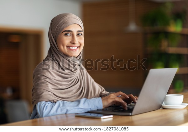 Cheerful muslim lady in hijab working online at cafe, using laptop and ... Cheerful muslim lady in hijab working online at cafe, using laptop and ...